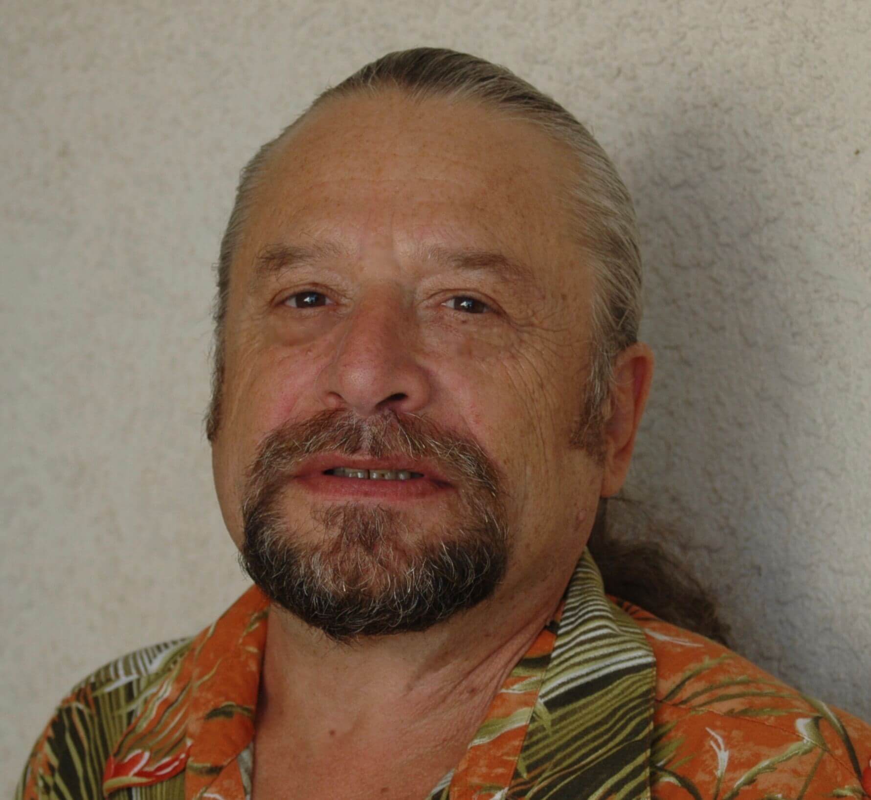 A man with long hair and beard wearing an orange shirt. - Myra Goldick